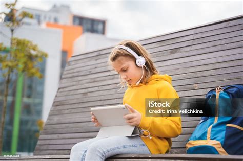 헤드폰에있는 소녀 태블릿이있는 헤드셋 공부 온라인 컨퍼런스 수업 교육 학교 마당 공원의 거리 벤치에서 원격 학습 배낭을 가진 여학생 채팅 친구와 대화 교사 1위에 대한 스톡