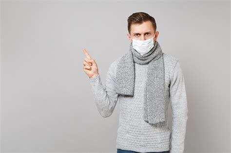 회색 스웨터에서 젊은 남자 스카프 메 마른 얼굴 마스크 포인팅 검지 손가락 옆으로 회색 벽 배경에 고립 건강 한 라이프 스타일