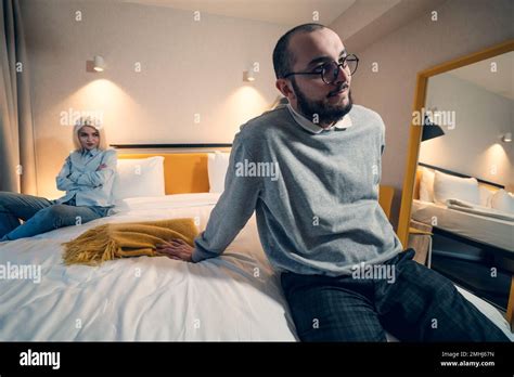 Worried Man Sit On Bed In Bedroom His Angry Wife Looks At Him Stock