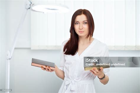 흰색 유니폼을 입은 매력적인 유능한 여성 미용사 미용사 전문의가 화장품 상자를 손에 들고 카메라로 현대 클리닉에서 실내에서