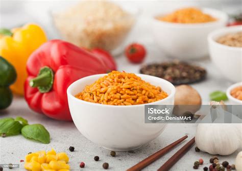 흰 그릇 접시에 야채와 버섯을 삶은 긴 곡물 바스마티 쌀이 있는 흰 그릇 접시에 막대기와 옥수수 마늘 바질이 있는 파프리카 페퍼가 있는 밝은 테이블 배경에 있습니다 건강한