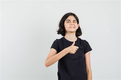 엄지 손가락을 보여주는 어린 소녀 검정 T 셔츠 전면보기에 눈을 윙크 무료 사진