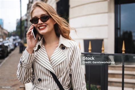 줄무늬 트렌치코트를 입은 젊은 예쁜 웃는 여자가 아늑한 도시 거리를 걷는 핸드폰에 행복하게 이야기합니다 16 17 살에 대한