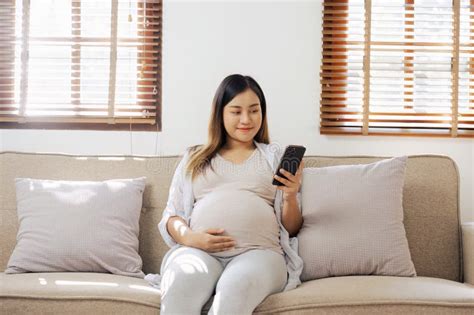 Happy Asian Pregnant Woman Using Smart Phone While Relaxing On Sofa In