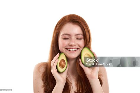 건강한 식단 젊고 행복한 빨간 머리 소녀의 초상화는 눈을 감고 흰색 배경에 서있는 동안 아보카도 두 조각을 들고 건강관리와