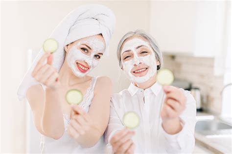 아침 스킨 케어 루틴 가족 스파 의 날 예쁜 선임 회색 머리 여자와 그녀의 젊은 매력적인 손녀 는 점토 얼굴 마스크를 만들고 오이 슬라이스와 부엌에서 카메라에 포즈 딸에