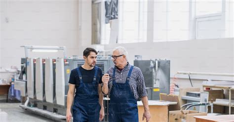 파란색 작업복을 입은 고위 및 젊은 인쇄소 노동자들이 현대 작업장을 건너 작업에 대해 논의합니다 사진 Unsplash의