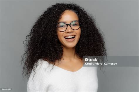 낙관적인 곱슬머리 여성의 솔직한 샷은 긍정적으로 웃고 기분이 좋고 건강한 피부를 가지고 있고 투명한 안경을 통해 보고 흰색