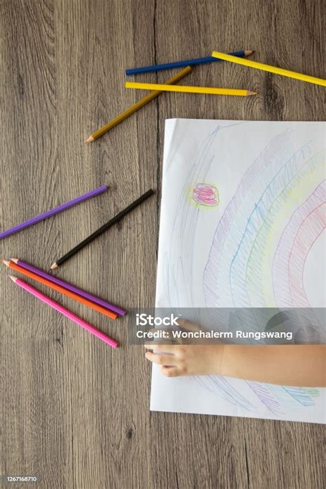위에서 찍은 사진은 미취학 아동이 나무 테이블에 놓인 흰색 종이에 컬러 연필로 그려진 다채로운 무지개 드로잉을 위해 창의력과
