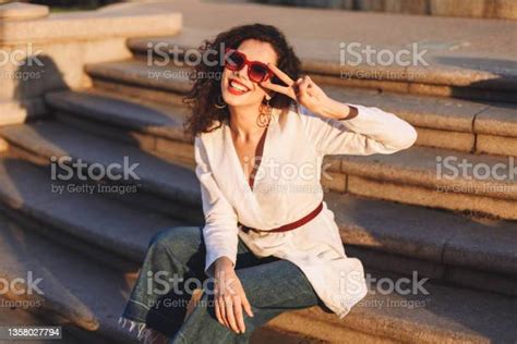 선글라스와 흰색 재킷을 입은 어두운 곱슬 머리를 가진 즐거운 아가씨가 거리의 계단에 앉아 두 손가락 제스처를 보여주면서 카메라를 즐겁게 바라보고 있습니다 20 24세에 대한