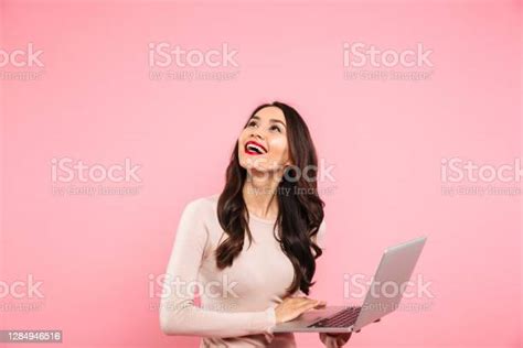 실버 노트북을 들고 핑크 배경에 고립 된 솔직한 미소로 카피 스페이스에서 위를 바라보는 캐주얼 스웨터에 갈색 머리 귀여운 여성의