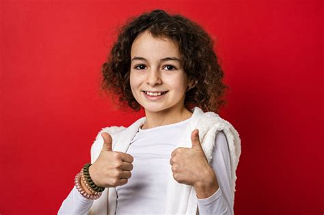 곱슬 머리 앞보기 초상화와 함께 10 살 된 작은 백인 소녀 10 살 은 행복하고 기쁨미소 복사 공간을 엄지 손가락을 들고 카메라를 찾고 빨간색 배경 앞에 서 10 11세에