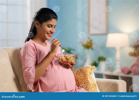 Happy Indian Pregnant Woman Eating Fruit Salad While Sitting On Sofa At