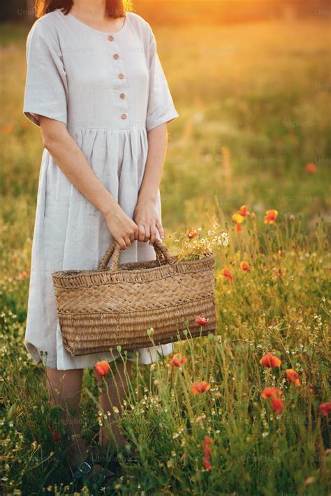 린넨 드레스를 입은 세련된 소녀는 일몰 빛 아래 초원에서 양귀비 꽃이 있는 소박한 짚 바구니를 들고 있습니다 여름 들판에서
