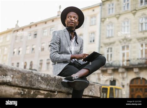 Focused African Man In Suit And Hat Sitting Outdoors And Using Digital