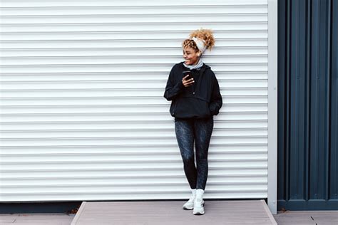 그녀의 전화와 이어버드를 사용하여 흰색 차고 문 앞에 서있는 행복한 아프리카 계 미국인 여성 먼 곳을 바라봄에 대한 스톡 사진