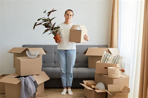 하얀 티셔츠와 청바지를 입고 새로운 아파트로 이사하고 골판지 상자에 개인 소지품을 들고 꽃 냄비에 심는 젊은 성인 백인 여성의 전체 길이 초상화 슬픔에 대한 스톡 사진 및