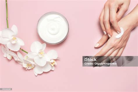 젊은 여자는 크림 바디 밀크와 함께 화장품 크림 로션 오픈 용기로 그녀의 손을 보습 핑크 배경에 화이트 Phalaenopsis 난초 꽃 플랫 누워 톱 뷰 미니멀리즘 스타일