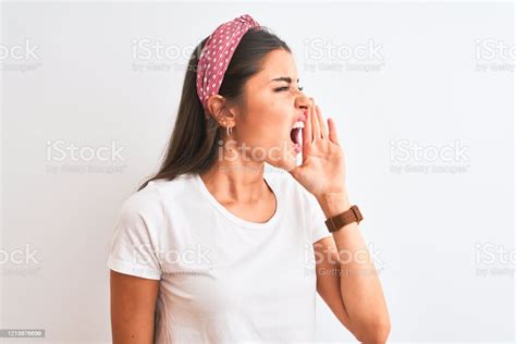 캐주얼 한 티셔츠와 디아뎀을 입고 고립 된 흰색 배경에 큰 소리로 외치고 입에 손을 대고 큰 소리로 비명을 지르는 젊은 아름다운