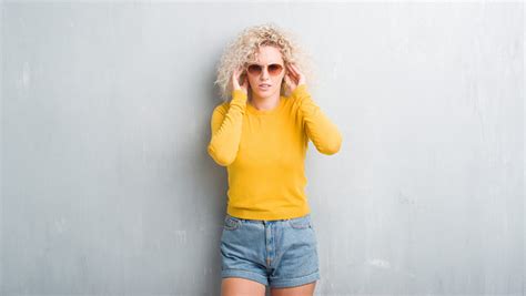 그런 지 회색 배경에 시끄러운 음악의 소음에 대 한 짜증이 식이 손가락으로 귀를 덮고 곱슬 머리를 가진 젊은 금발 여자 청각