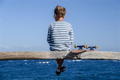 바다를 보고 벤치에 앉아 안경을 가진 잘생긴 아이 십 대 휴식과 그의 스케이트와 야외를 즐기는 세련 된 작은 소년은 스케이트 보드와 함께 바다 근처 아름 다운 여름 날을