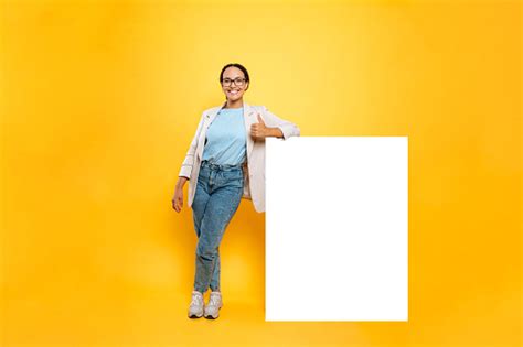 긍정적인 브라질 여성의 전체 길이 사진 프레젠테이션 발표 또는 광고를 위한 정사각형 흰색 모형 포스터 근처에 서 있고 엄지손가락을 치켜세우는 제스처 미소 고립된 노란색