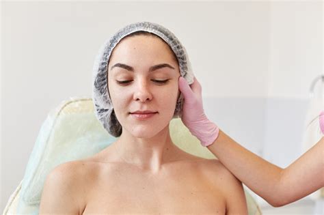 화장품 살롱에서 직장에서 분홍색 의료 장갑의 얼굴 없는 미용 학자의 이미지는 매력적인 여 자가 일회용 캡에 Couh 앉아 아래로 보고 검사 합니다 미용 및 Aethetic