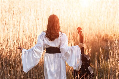 흰색 중세 드레스와 어쿠스틱 기타를 들고 일몰 조명 필드를 통해 걷는 벨트에 빨간 머리를 가진 날씬한 젊은 여성의 다시 보기 렌즈 플레어 가을에 대한 스톡 사진 및 기타