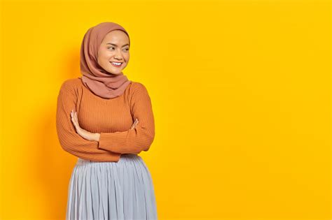 갈색 스웨터와 히잡 을 입은 아름다운 미소 짓는 아시아 여성이 팔 가슴을 가로 질러 노란색 배경 위에 고립 된 복사 공간을