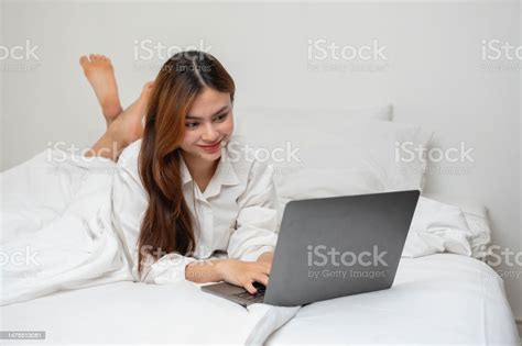 하얀 잠옷을 입은 여자가 주말 아침에 일어나 휴식을 취하고 노트북 휴대폰을 가지고 놀며 하얀 침실 안의 유리에서 빵을 먹고 차를 마신다 아침 휴가 개념입니다 갈색 머리에