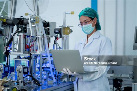 ガウンと衛生マスクを持つ工場労働者の女性は工場の職場でマシンを制御するためにラップトップを保持します 実験室のストックフォトや画像を多数ご