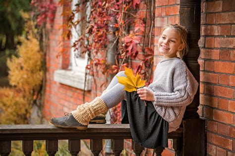 따뜻한 스웨터를 입은 한 소녀가 가을의 자연 펜스 평화로운 배경에 대해 노란 단풍 잎이 달린 오래된 오두막의 베란다의 나무 울타리에 앉아 있습니다 8 9 살에 대한 스톡