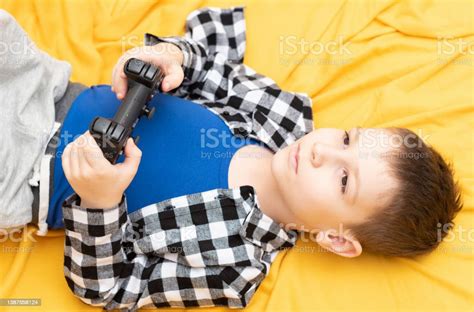 체크 셔츠를 입은 아이는 비디오 게임을 하는 손에 검은 조이스틱을 들고 소파에 누워 있었다 홈 컨셉에서 비디오 게임 재생 6 7 살에 대한 스톡 사진 및 기타 이미지