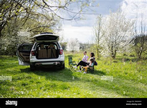 Mother With Two Daughters Sit On Chair Against Car Open Trunk On Picnic