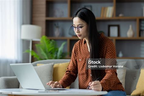 노트북에서 집에서 일하고 펜과 종이로 작업에 집중하고 편안한 소파에 앉아 세련된 거실 환경에서 원격 근무 라이프 스타일을 보여주는 젊은 아시아 여성 교육에 대한 스톡 사진