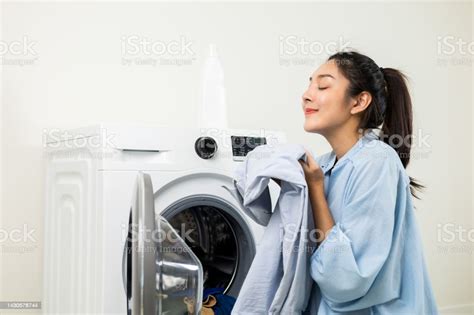 행복한 주부는 세탁기 앞쪽을 열고 집에서 씻은 세탁물을 받기 위해 미소 지었다 연화제와 세탁기에서 씻은 향기로운 깨끗한 옷 냄새를 맡는 여성 섬유유연제에 대한 스톡 사진 및