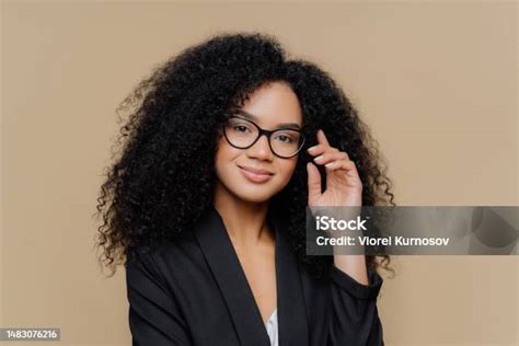 차분한 만족 아프로 미국 여성의 초상화를 닫아 부시 컬 헤어 스타일로 손을 들고 건강한 피부를 가지고 안경을 착용하고 갈색 벽에 고립 된 검은 우아한 의상을 착용합니다 얼굴