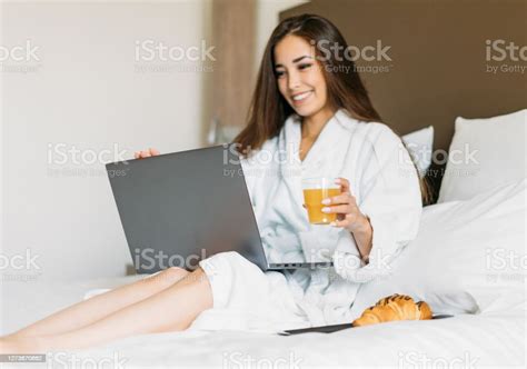 호텔 방 침대에서 아침 크루아상과 오렌지 주스를 가지고 노트북을 사용하여 흰색 가운에 긴 머리를 가진 젊은 아시아 소녀 20