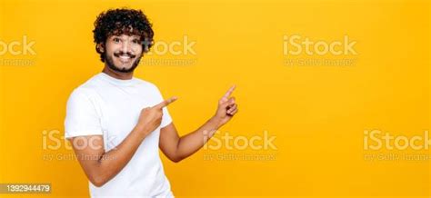 기본적인 흰색 티셔츠를 입은 긍정적 인 인도 또는 아라비안 녀석은 카메라를보고 빈 공간에서 손가락을 옆으로 가리키며 고립 된