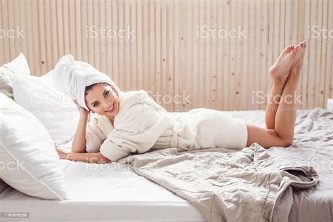 흰색 목욕 가운과 그녀의 머리에 수건으로 꽤 행복한 젊은 여자는 아늑한 집에서 침대에 누워있다 아름다운 소녀는 아침에 침대에서