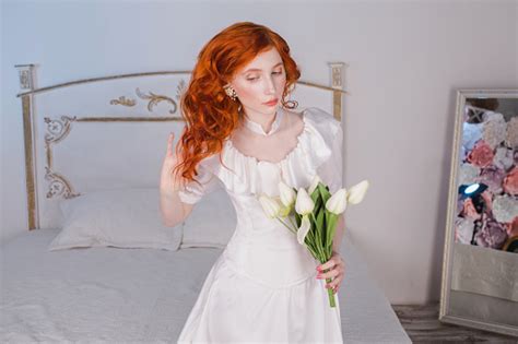 귀에 하얀 진주 귀걸이와 흰색 빈티지 웨딩 드레스에 긴 붉은 곱슬 머리 여자 창백한 피부 파란 눈 손에 흰색 튤립으로 침실에 특이 한 모양으로 빨간 머리 소녀 결혼식에 대한