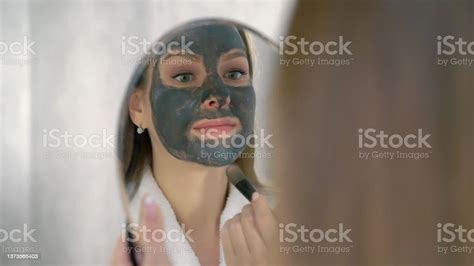 젊은 여성은 얼굴의 피부를 돌봅니다 소녀는 회색 점토 화장품 마스크를 착용 거울에 대한 스톡 사진 및 기타 이미지 거울