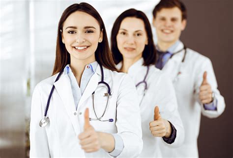 젊은 쾌활한 의사 그룹이 병원 사무실에서 엄지 손가락을 치켜 들고 팀으로 서서 환자를 도울 준비가되어 있습니다 의료 지원건강 관리 보험 20 24세에 대한 스톡 사진 및