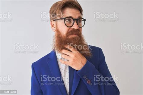 젊은 빨간 머리 아일랜드어 사업가 는 질문 펜시브 표현에 대해 생각 턱에 손으로 고립 된 흰색 배경 위에 양복과 안경을 착용 사려 깊은 얼굴로 미소 의심 개념 경영자에 대한