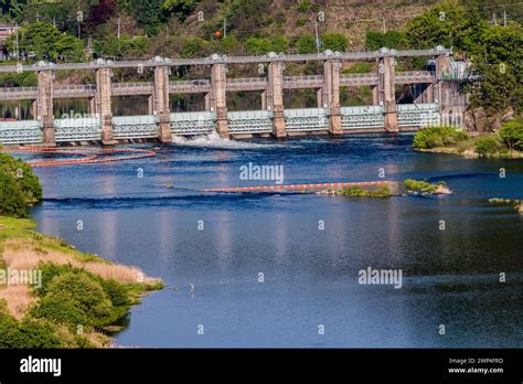 Landscape Of Dam Releasing Water Through One Floodgate Into River With Blue Water Located Near