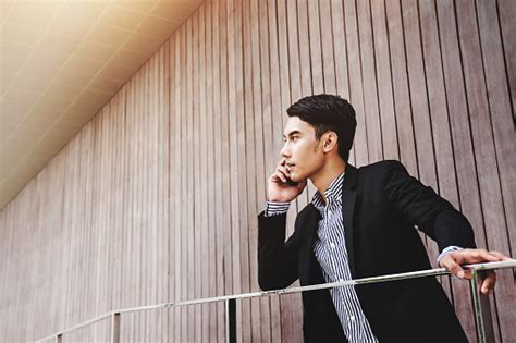 나무 벽에 심각 하 게 얼굴을 가진 똑똑한 전화를 통해 젊은 아시아 사업가 이야기 검정 머리에 대한 스톡 사진 및 기타 이미지 검정 머리 기업 재무와 산업 남자