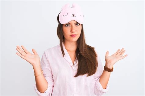 잠옷과 수면 마스크를 입고 고립 된 흰색 배경 에 서있는 젊은 아름다운 소녀는 팔과 손을 들고 단서와 혼란스러운 표정을 하고 있습니다 의심 개념 갈색 머리에 대한 스톡 사진