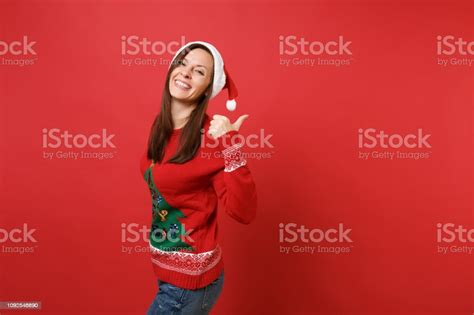 메리 산타 소녀 니트 스웨터 크리스마스 모자 포인팅 엄지손가락 옆으로 붉은 벽 배경에 고립의 측면 볼 수 있습니다 행복 한 새 해 2019 축 하 휴일 파티 개념입니다 복사