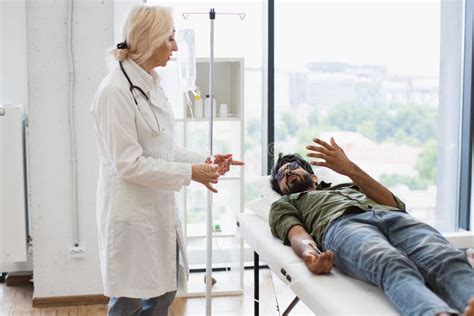 Bearded Young Man Receiving Chemotherapy Looking At Nurse In Lab Coat