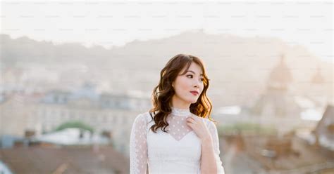 하얀 드레스를 입은 우아한 아시아 여성이 옥상 테라스에 서서 일몰에 옛 유럽 도시의 도시 배경 스카이라인 전망을 보고 있다 오래 된 도시 전망의 배경에 포즈를 취하는 중국